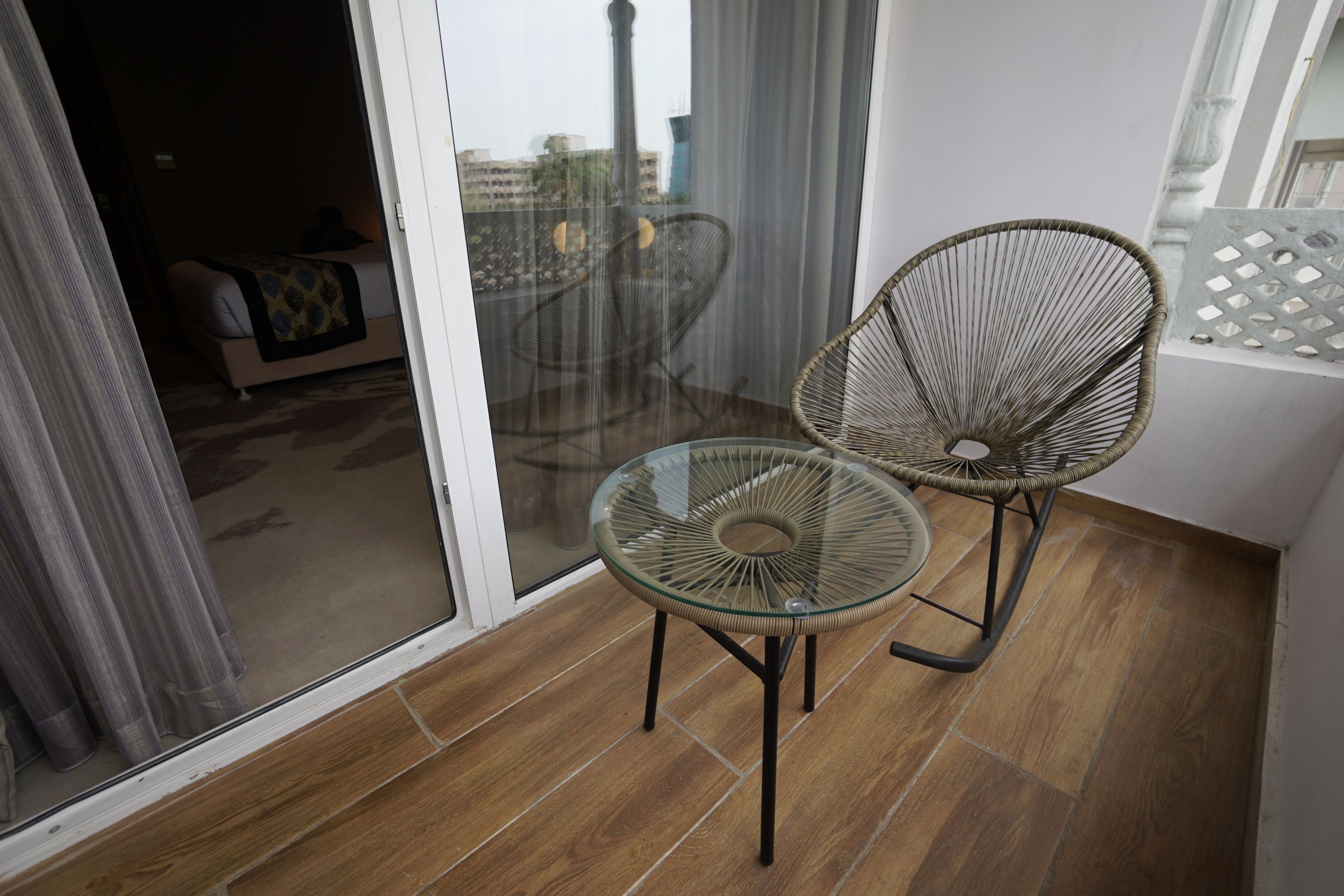 Uma varanda com mesa de vidro e duas cadeiras de vime, porta de correr com vidro e vista para edifícios ao fundo.