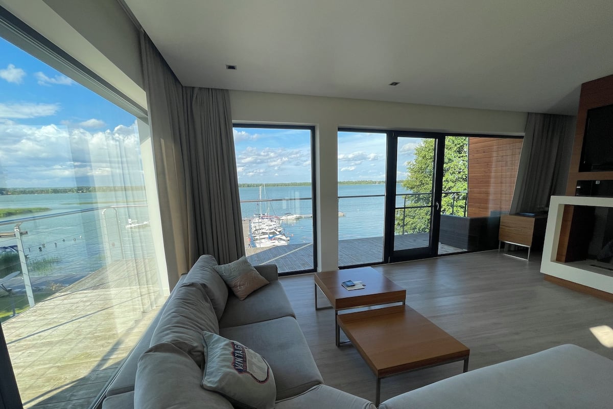 Uma sala de estar moderna com um sofá grande, uma mesa de centro e uma televisão. Possui janelas amplas com vista para um lago e um píer.