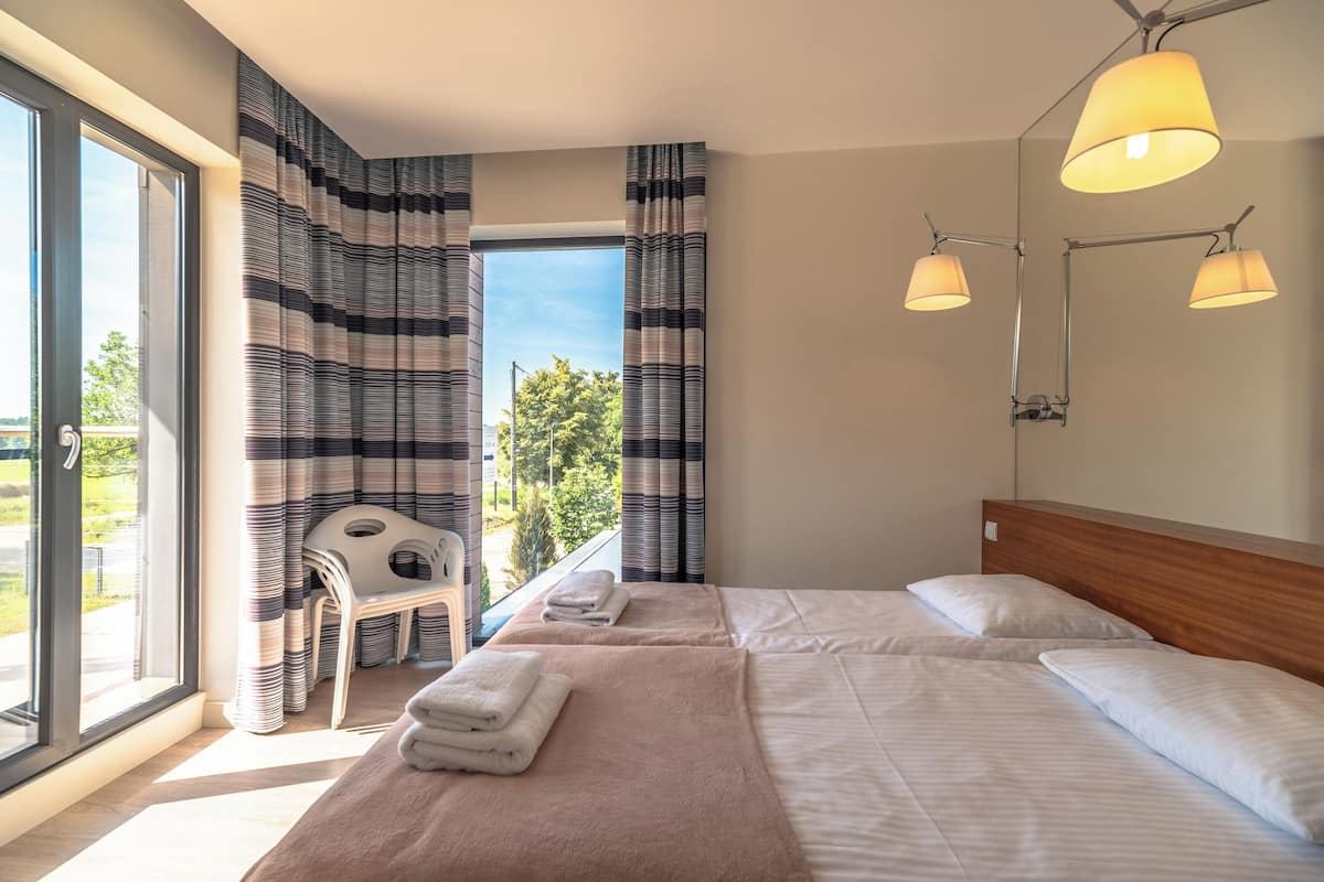 Quarto de hotel com duas camas, uma janela grande com cortinas, uma cadeira e uma varanda.