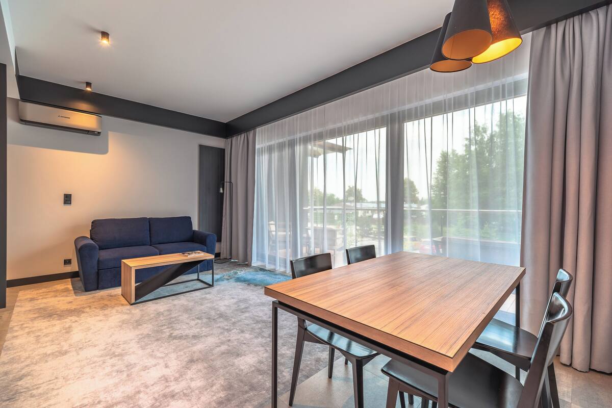 Uma sala de estar moderna com um sofá, uma mesa de jantar de madeira e cadeiras. Há uma janela grande com cortinas leves e vista para o verde lá fora.