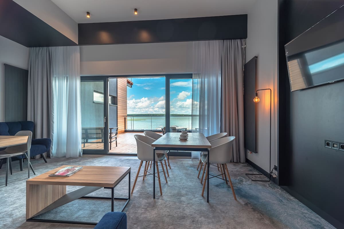 Quarto de hotel moderno com uma grande porta de vidro deslizante que dá para uma varanda com vista para o mar. Há uma mesa de jantar com cadeiras, um sofá e uma televisão fixada na parede.