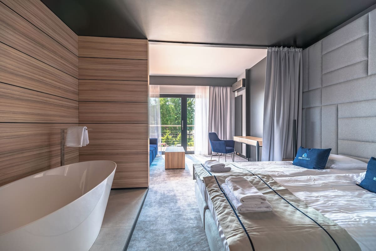 Quarto de hotel moderno com uma banheira grande, uma cama com toalhas dobradas com cuidado, uma pequena escrivaninha e vista para o exterior.