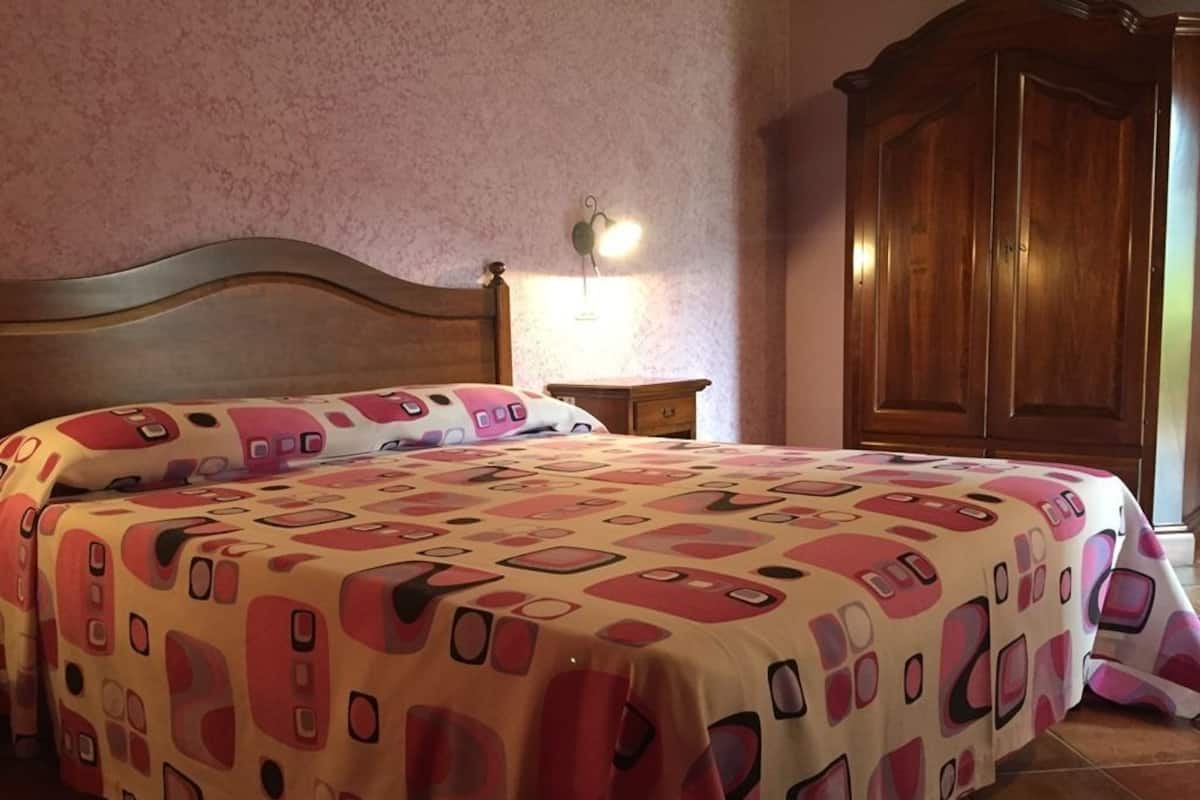 Quarto com cama coberta por colcha estampada, cabeceira de madeira, mesa de cabeceira com abajur e guarda-roupa.