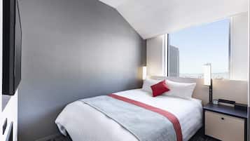 Quarto de hotel moderno com uma cama grande, uma mesa de cabeceira, televisão e vista para a cidade pela janela.