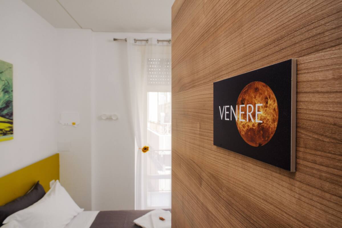 Quarto de hotel com cama, parede de madeira e um quadro com a lua, exibindo a palavra "VENERE".
