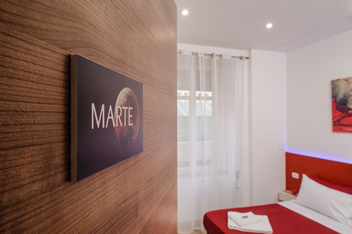Quarto de hotel com parede de destaque em madeira, cama com roupa de cama vermelha, mesa de cabeceira branca e janela com cortinas leves.