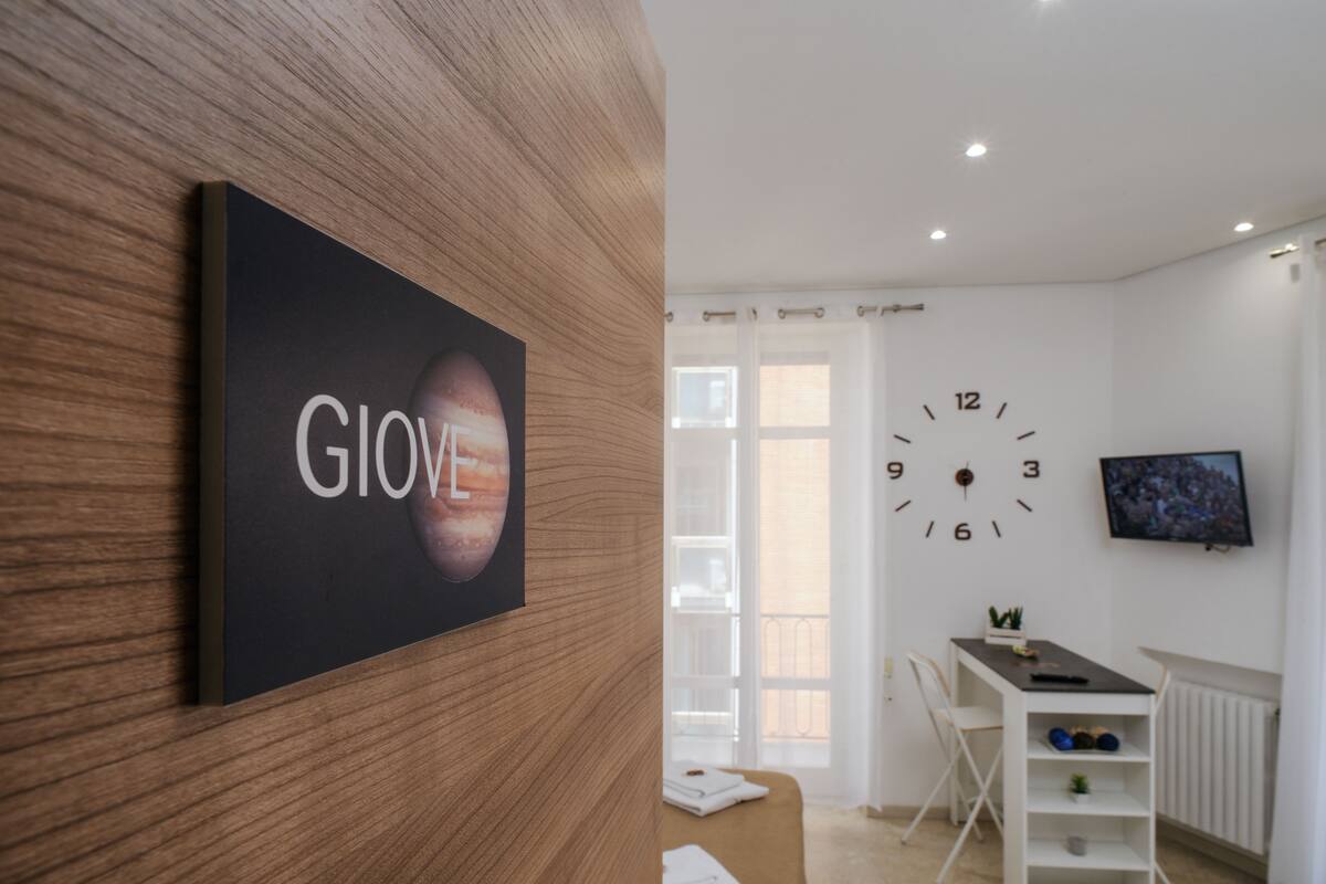 Quarto com parede de madeira, relógio, televisão, escrivaninha e cadeira.