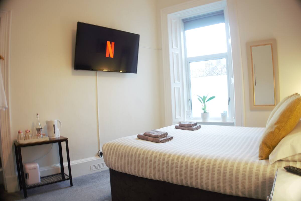 Quarto de hotel com uma cama, televisão na parede, uma mesinha com chaleira e uma janela com planta.