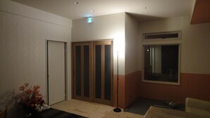 Quarto de hotel com chão de madeira, uma cama, uma escrivaninha, uma cadeira, uma janela e uma porta.
