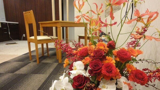 Um quarto com piso coberto de carpete, uma cadeira de madeira e um arranjo floral sobre uma mesa.