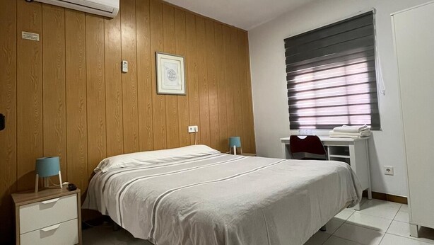 Quarto com cama, mesas de cabeceira, escrivaninha, janela com persianas, ar condicionado e quadro na parede.