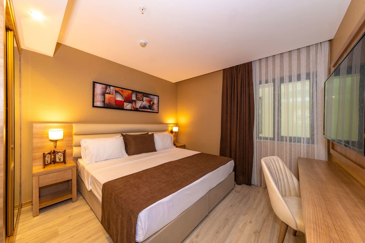 Quarto de hotel com uma cama grande, luminárias de cabeceira, uma escrivaninha, uma cadeira e um quadro na parede.