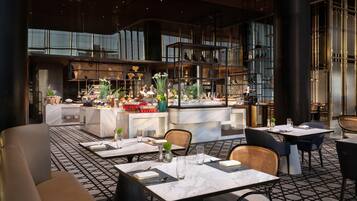 Um restaurante moderno com mesas de mármore, cadeiras de vime e um bar central com diversas opções de comida e bebida.