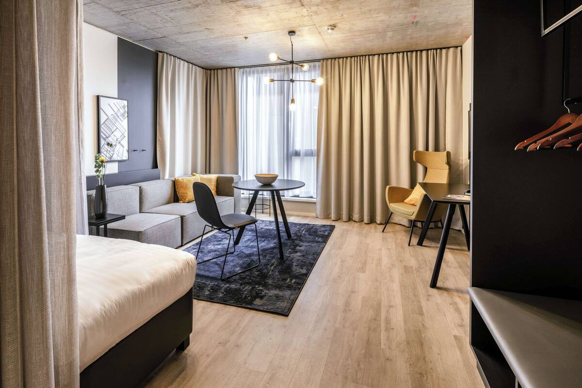 Quarto de hotel moderno com cama, sofá, cadeiras, uma pequena mesa e um cabideiro.