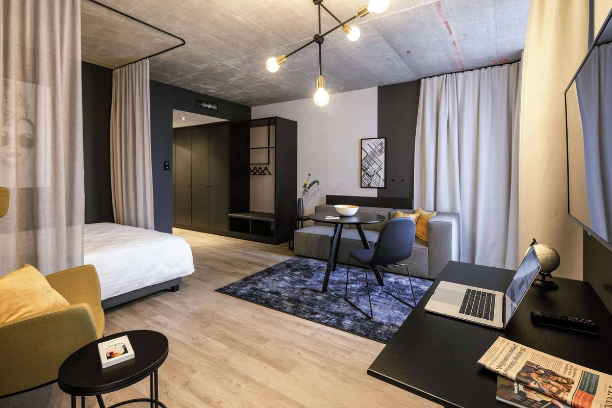 Quarto de hotel moderno com cama, escrivaninha com laptop, mesa pequena e sofá.