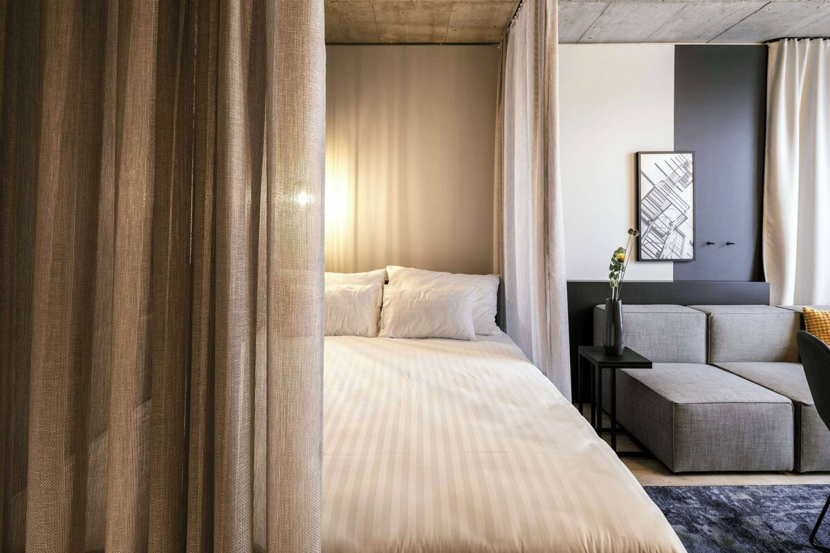 Quarto de hotel moderno com uma cama grande, um sofá cinza e uma pequena mesa com um vaso.