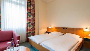 Quarto de hotel com duas camas, uma poltrona vermelha, uma mesa pequena com um jogo de copos e uma janela com cortinas.