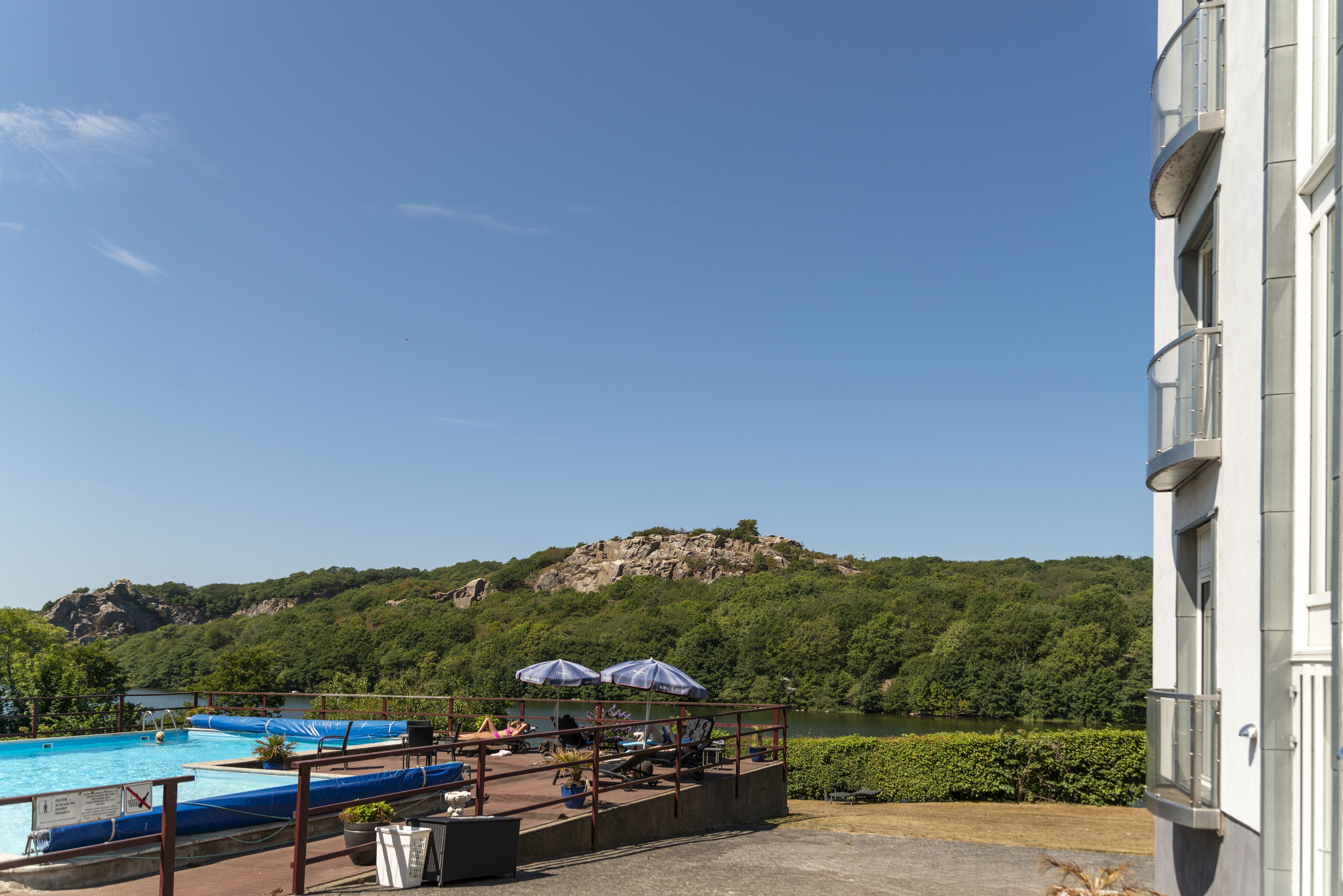 Uma área de piscina com espreguiçadeiras e guarda-sóis, vista para um lago e um edifício com varanda com vista para a paisagem.