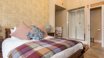 Quarto com uma cama grande, cabeceira de madeira e manta xadrez. Apresenta uma mesinha de cabeceira com abajur e uma pequena mesa com um vaso. Um banheiro com chuveiro de vidro e porta-toalhas é visível ao fundo.