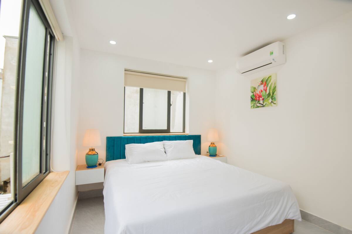 Quarto com cama, duas mesas de cabeceira, janela, aparelho de ar condicionado e um quadro na parede.