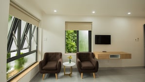 Quarto moderno com duas poltronas marrons, uma mesa redonda pequena com um vaso, uma janela grande com vista para o verde, uma TV integrada e uma escrivaninha de madeira com uma gaveta.