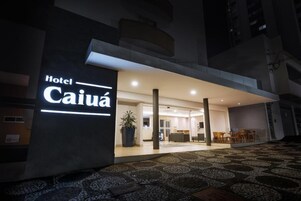 Entrada de hotel com design moderno, um letreiro grande e iluminado, e uma área de recepção com assentos.