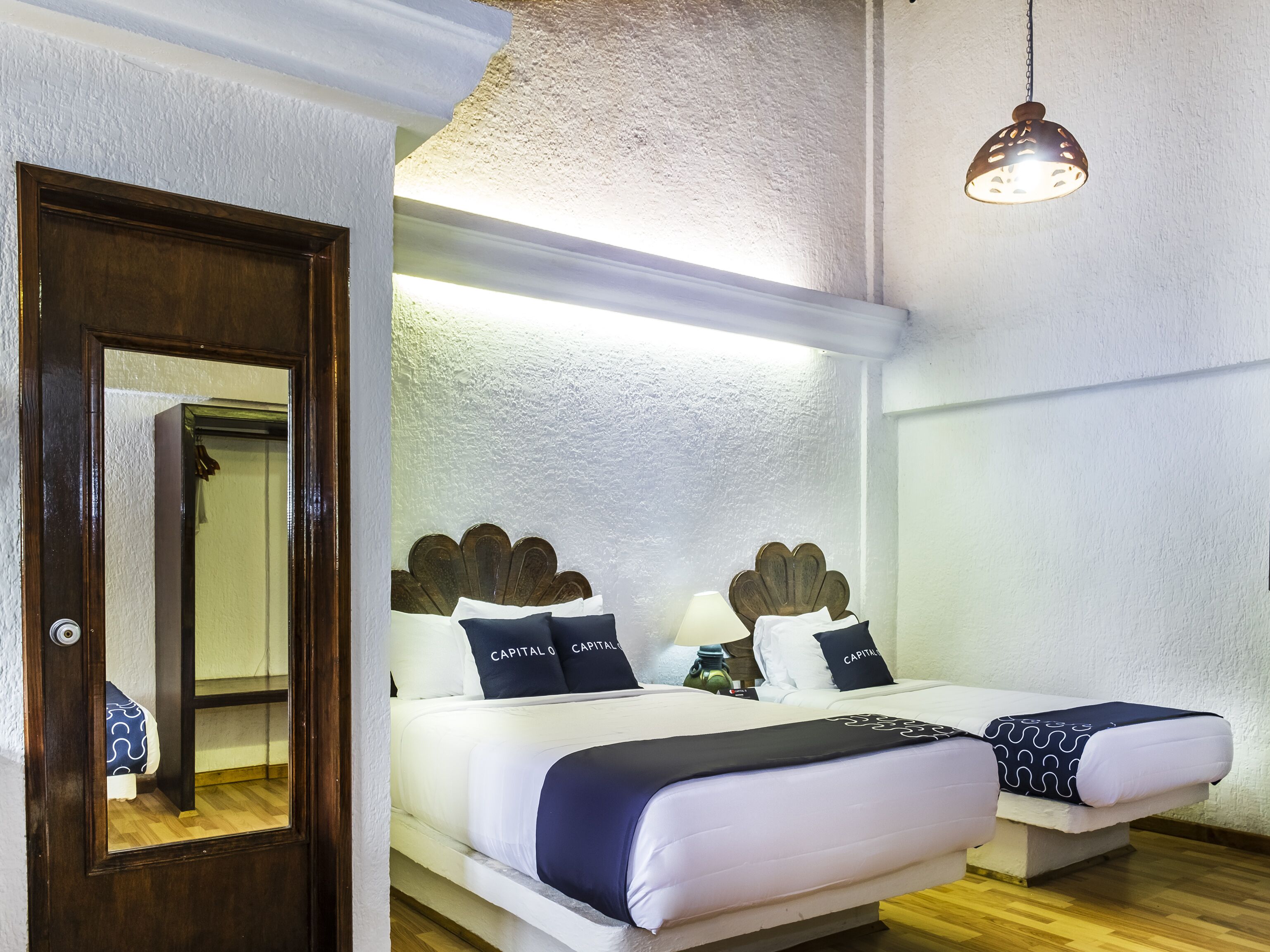 Quarto de hotel com duas camas, cada uma com cabeceira, roupa de cama branca e manta azul escura. Há uma porta de madeira e um espelho na parede.