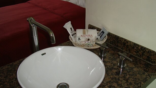 Uma pia branca com torneira cromada, uma cama vermelha e um cesto com amenities sobre um balcão de granito.