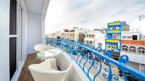 Uma varanda com móveis de vime branco, guarda-corpo azul e vista para edifícios e montanhas ao fundo.