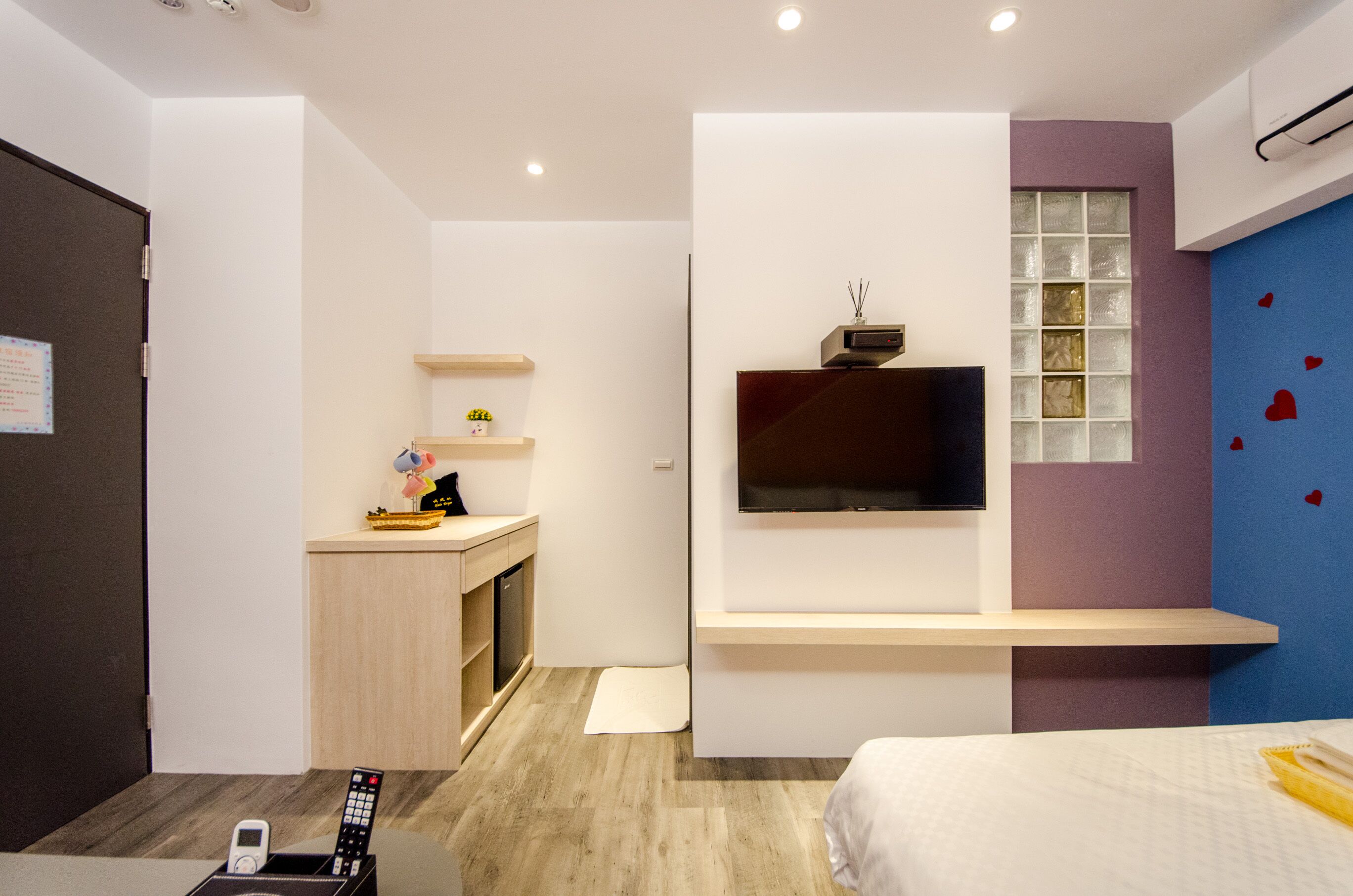 Quarto de hotel moderno com televisão de tela plana na parede, uma escrivaninha pequena com abajur e uma cama com edredom branco.