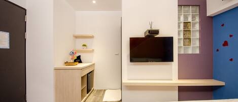 Quarto de hotel moderno com televisão de tela plana na parede, uma escrivaninha pequena com abajur e uma cama com edredom branco.