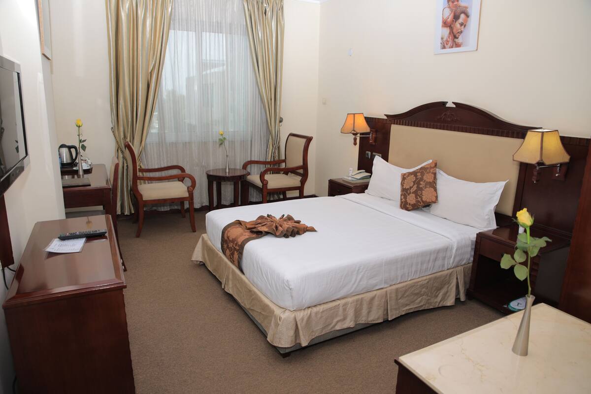 Quarto de hotel bem arrumado, com uma cama grande, duas mesas de cabeceira, uma escrivaninha e uma cadeira.