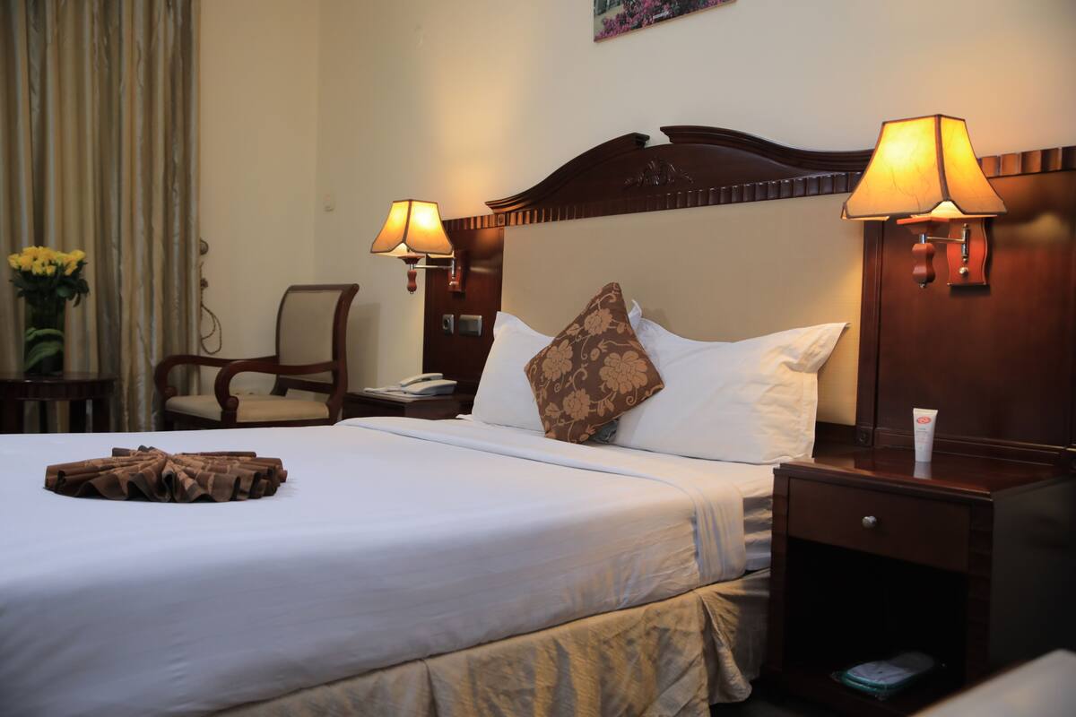 Quarto de hotel com uma cama grande, duas luminárias de cabeceira, uma cabeceira de madeira, uma mesa de cabeceira com luminária, uma cadeira e um vaso com flores.