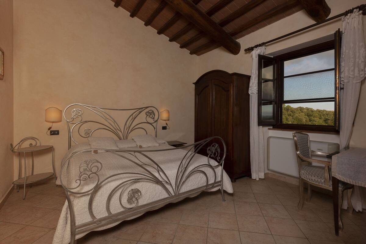 Quarto com estrutura de cama de metal, guarda-roupa de madeira, mesa pequena com cadeira e janela com vista para o verde.