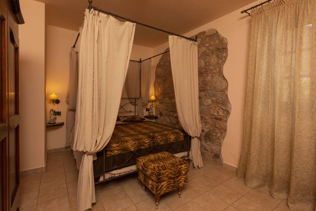 Quarto com cama de dossel, parede de destaque em pedra e um pequeno banco de madeira.