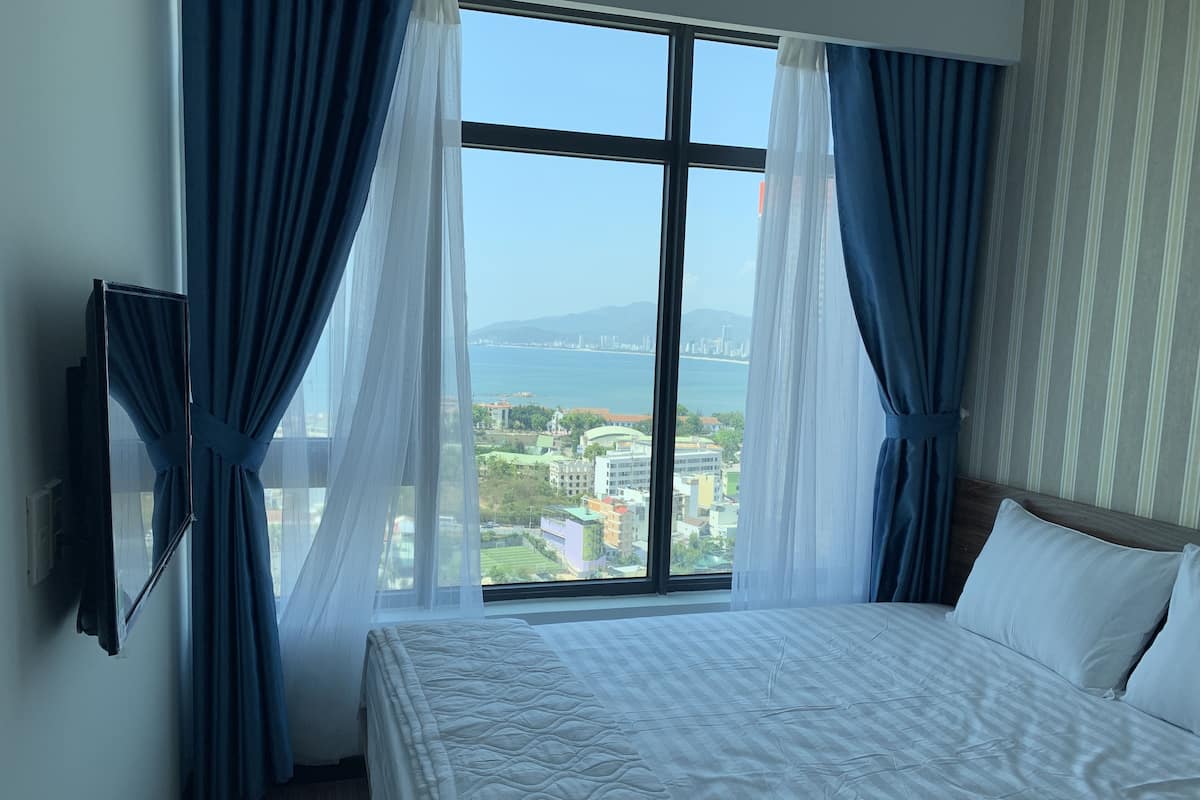 Quarto de hotel com uma cama grande, televisão na parede e vista para a cidade através de uma janela ampla.