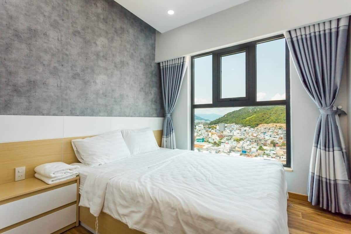 Quarto de hotel com uma cama grande, cabeceira de madeira, mesa de cabeceira e vista para a cidade através de uma janela com cortinas.