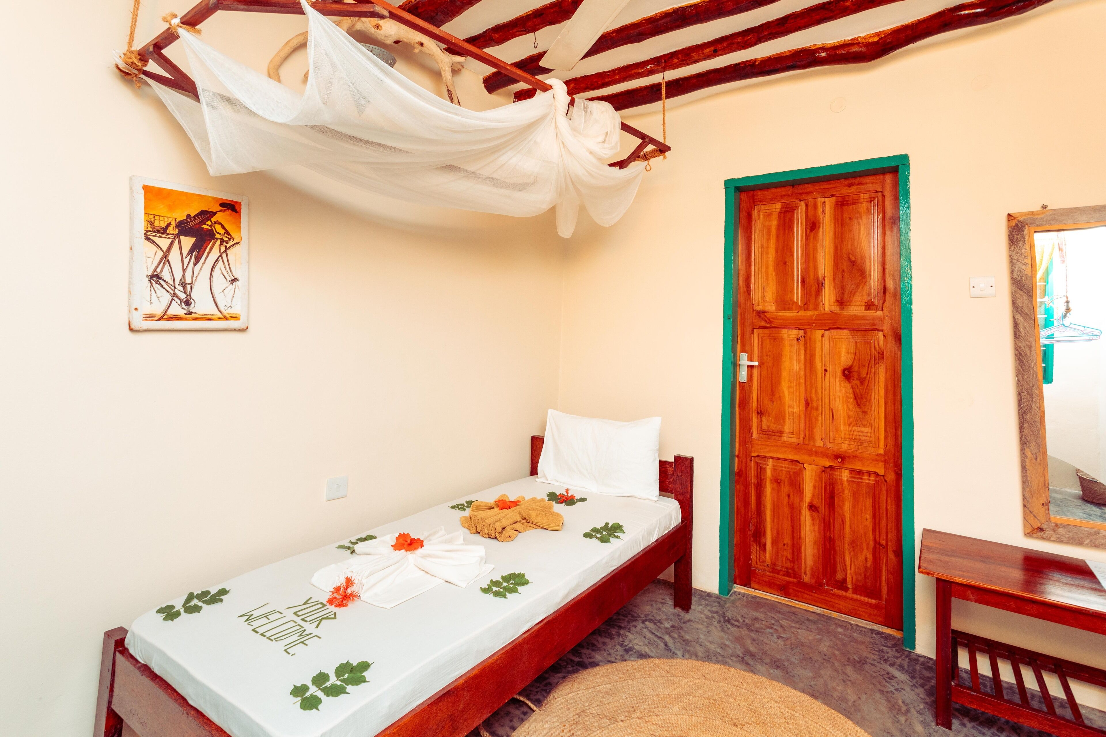 Uma cama de solteiro com lençol branco, um tapete de boas-vindas, uma porta de madeira e um pequeno banco de madeira.