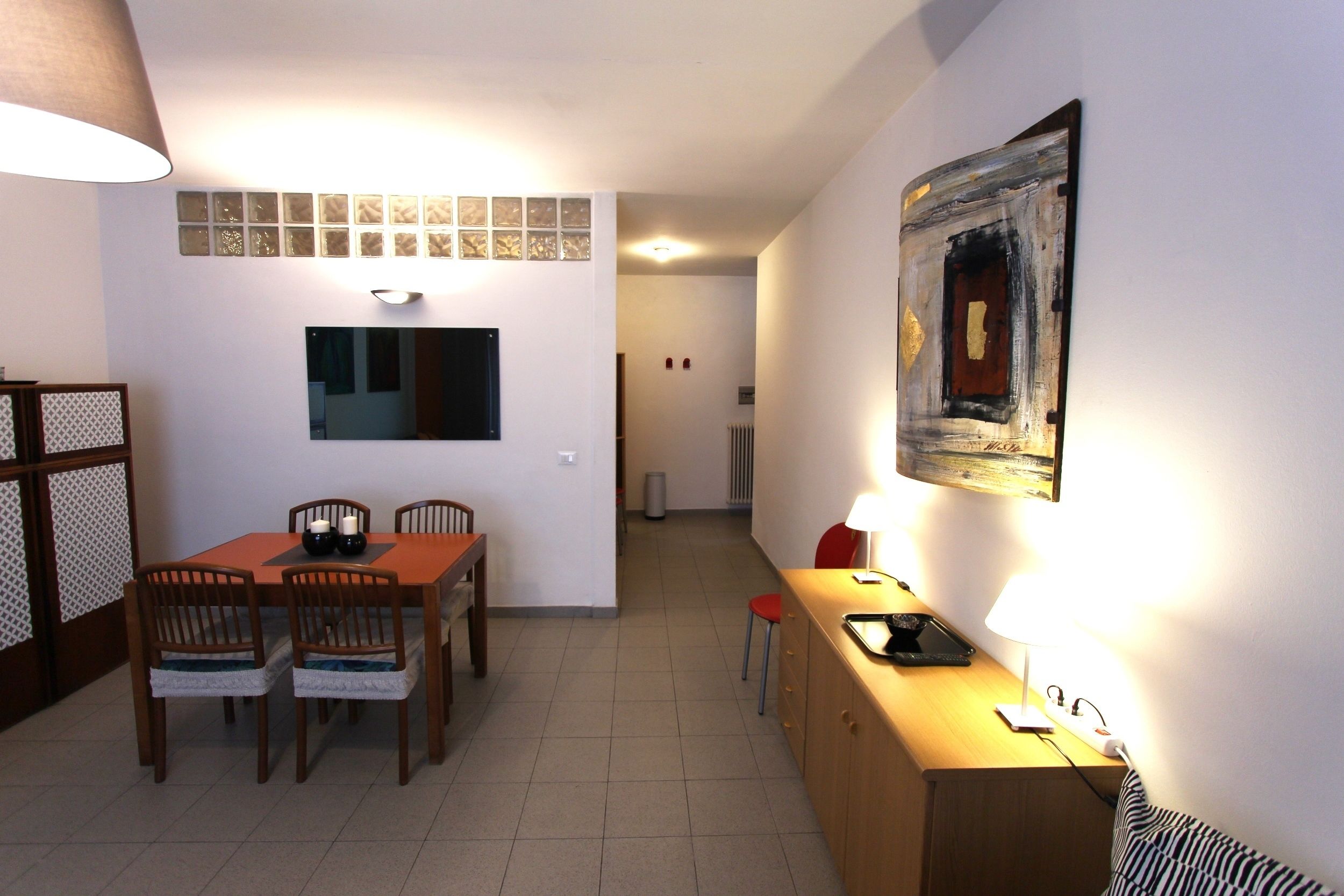 Uma área de refeições com uma mesa e cadeiras de madeira, um banquinho pequeno, um armário com uma luminária e um quadro fixado na parede.