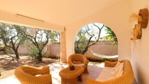 Um pátio ensolarado com móveis de vime, uma rede e vista para árvores e um muro.