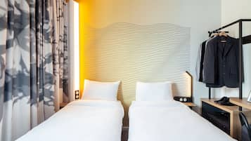 Quarto de hotel com duas camas, cabeceira com design ondulado, mesa de cabeceira com abajur e um cabideiro com um terno.