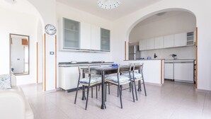 Uma cozinha moderna com armários brancos, uma área de jantar com mesa e cadeiras redondas, e um relógio visível na parede.