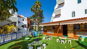 Uma área de recreação infantil com grama sintética, mesas, cadeiras e um parquinho de madeira.