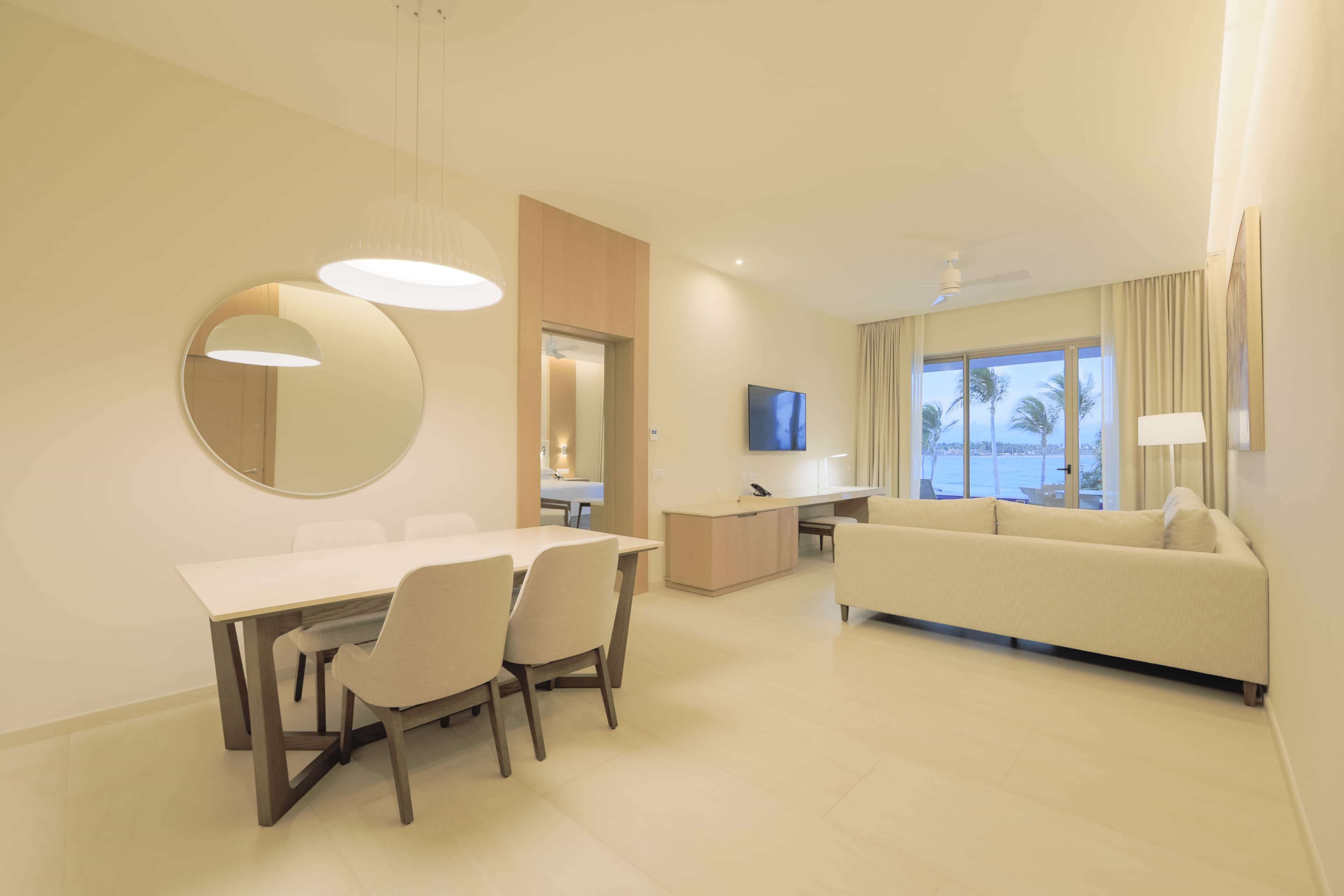 Quarto de hotel moderno com área de refeições, sofá, TV e vista para palmeiras.