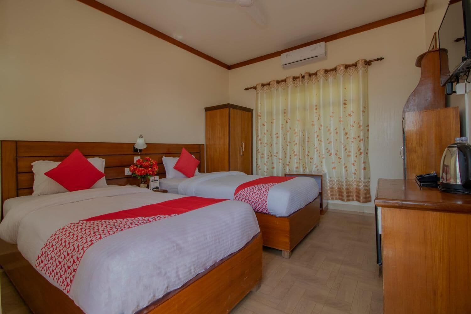 Quarto de hotel com duas camas, cabeceiras de madeira, cômoda de madeira, unidade de ar condicionado e cortina com padrão floral.