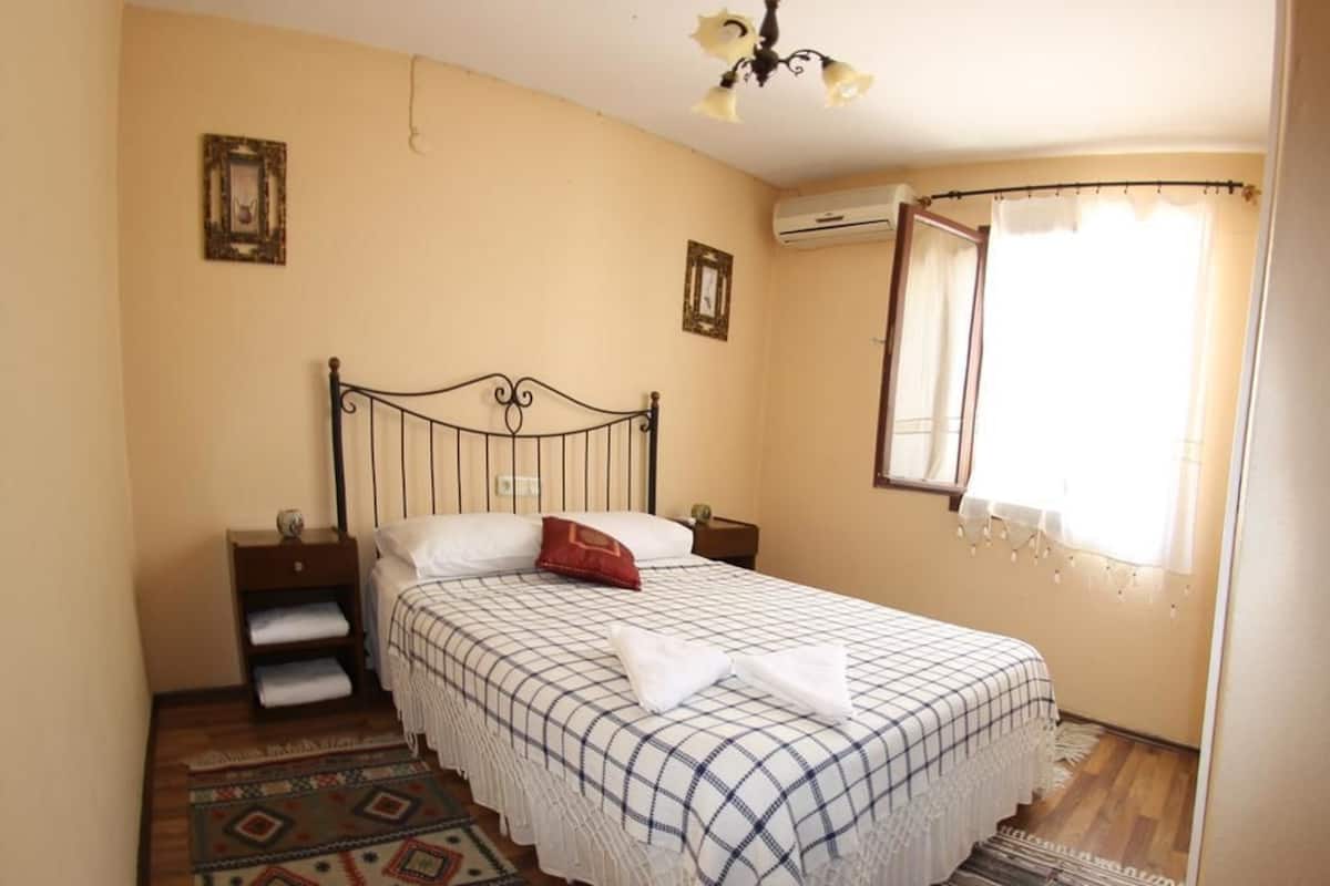Quarto com cama, mesas de cabeceira, janela com cortinas e um tapete estampado.