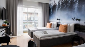 Quarto de hotel moderno com duas camas, uma escrivaninha com televisão e uma janela grande com cortinas.