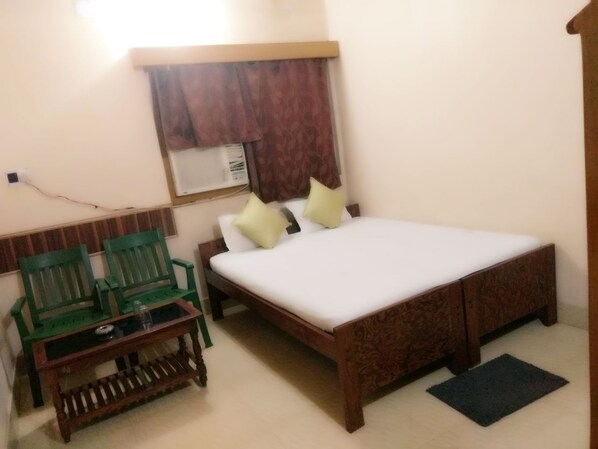 Quarto com uma cama, duas cadeiras verdes, uma mesa pequena, uma janela com cortinas e um ar condicionado fixado na parede.