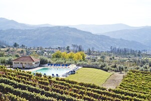 Uma área de piscina com espreguiçadeiras, um edifício de pedra e vinhedos em primeiro plano, com montanhas ao fundo.