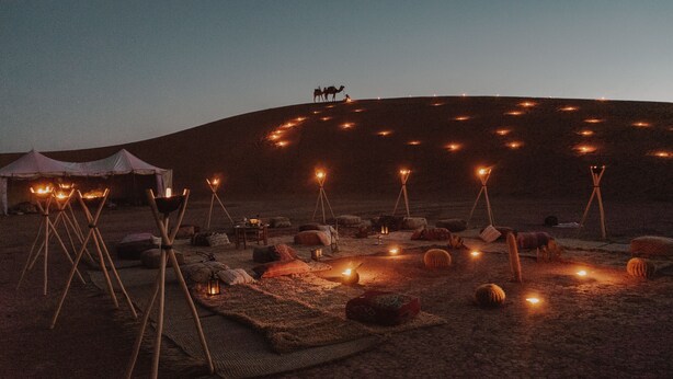 Acampamento no deserto ao entardecer, com tendas, lanternas e camelos.